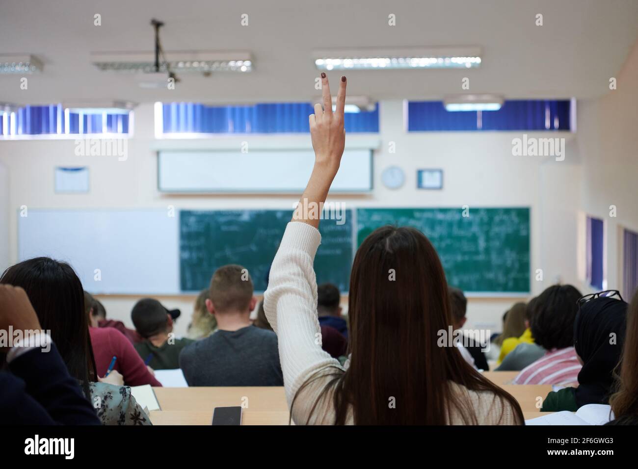 Rear view of female student sitting in the class and raising hand up to ...