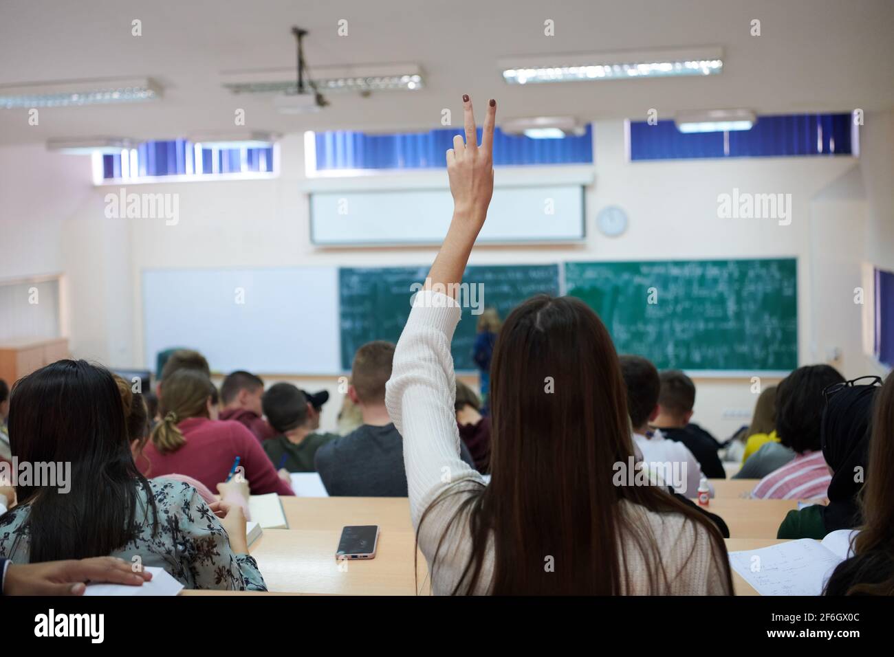 Rear view of female student sitting in the class and raising hand up to ...
