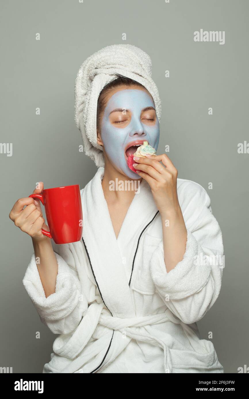 Happy young woman in cosmetic face mask eating breakfast and drinking ...