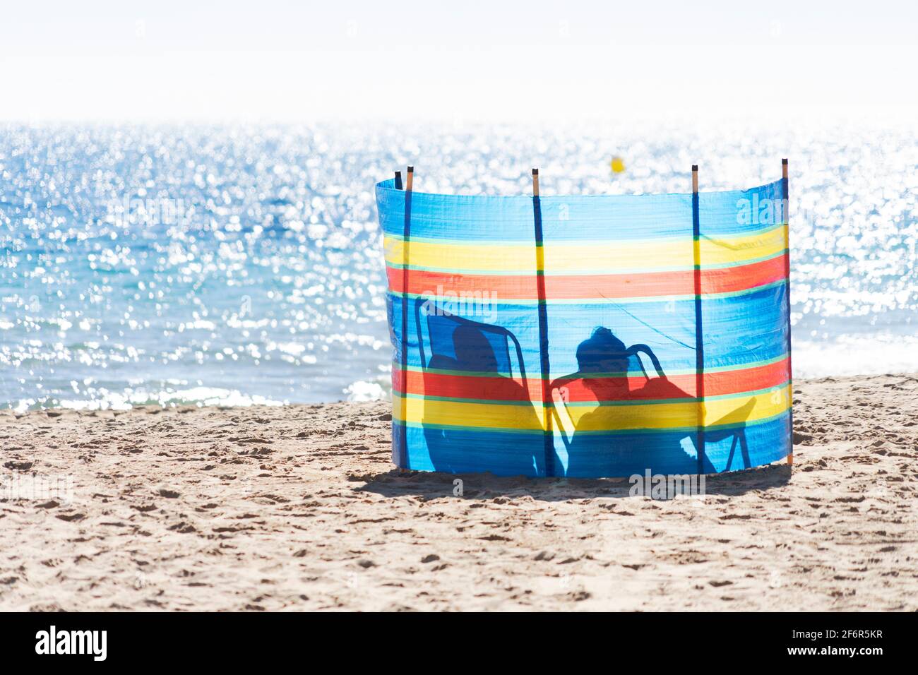 Two people relaxing behind colorful striped windscreen on the beach ...