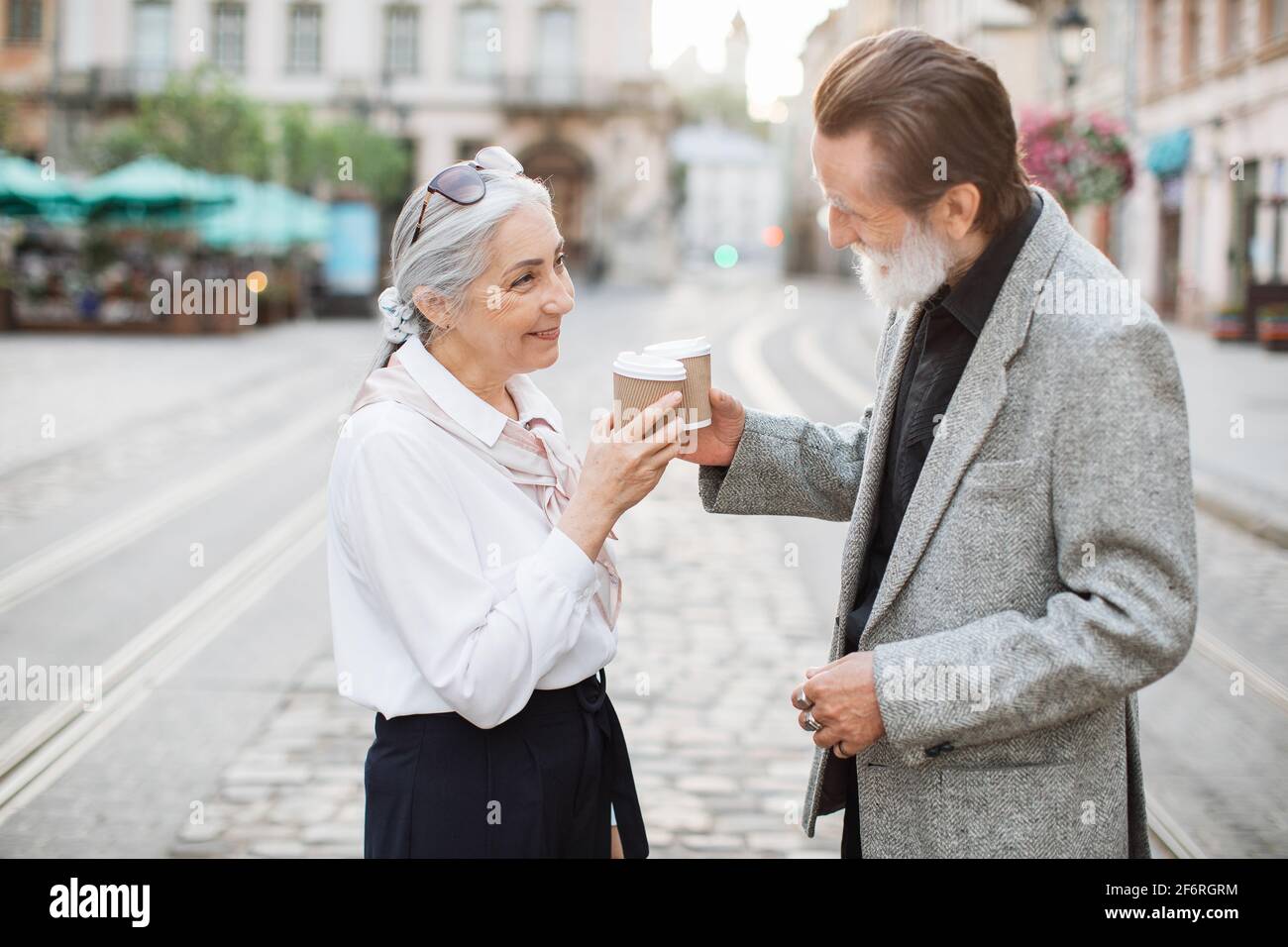 Side view of smiling aged couple in stylish outfits toasting with ...
