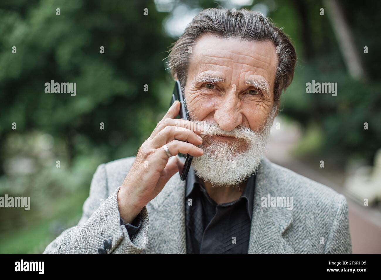 Mature bearded man standing at green park and talking on mobile. Confident stylish pensioner ...