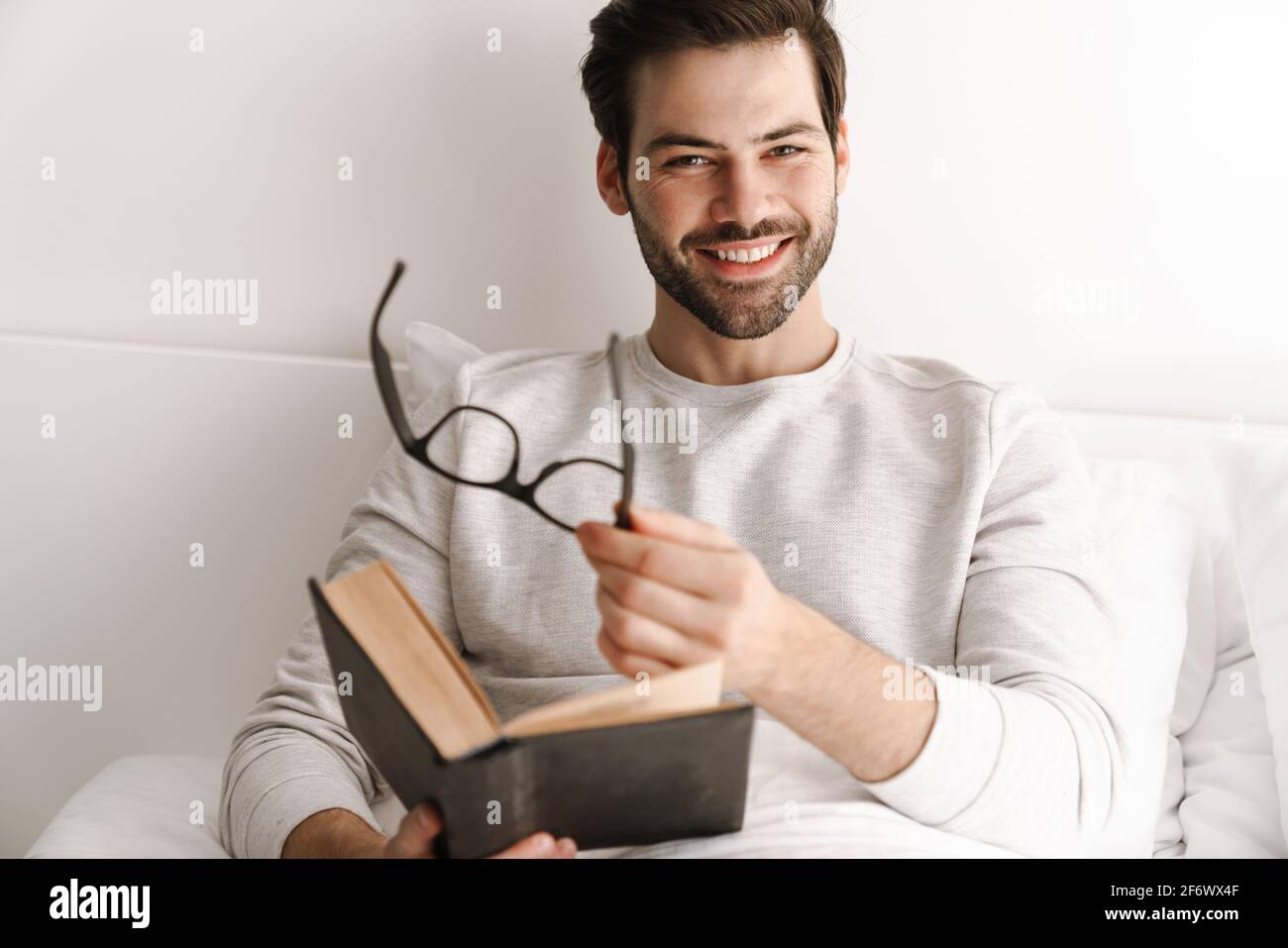 Happy young man smiling and reading book while resting in bed at home ...