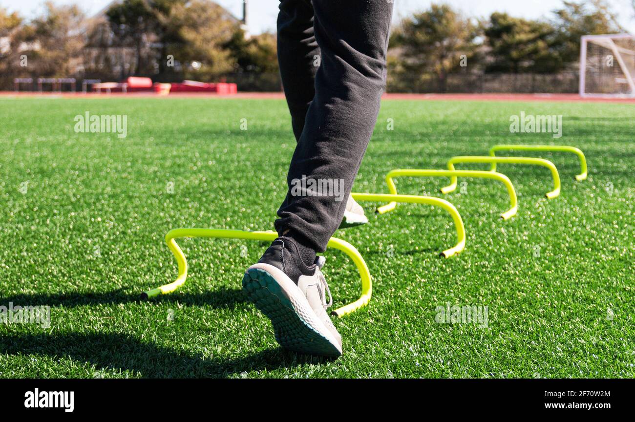 The feet of an athlete stepping over yellow mini banana hurdles set up aon a green turf field ...