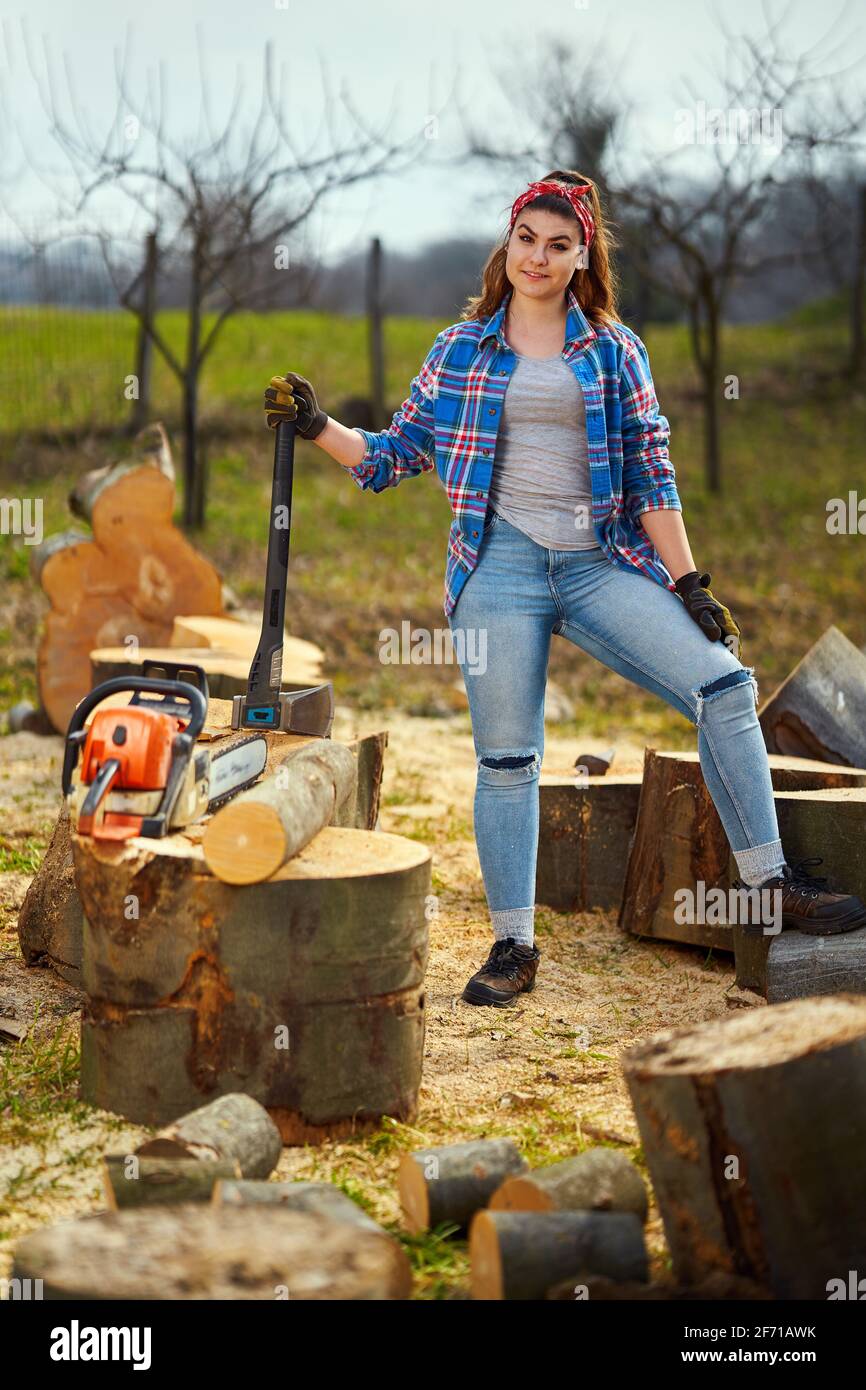 Strong woman woodcutter with her ax and her chainsaw amongst huge sawn ...