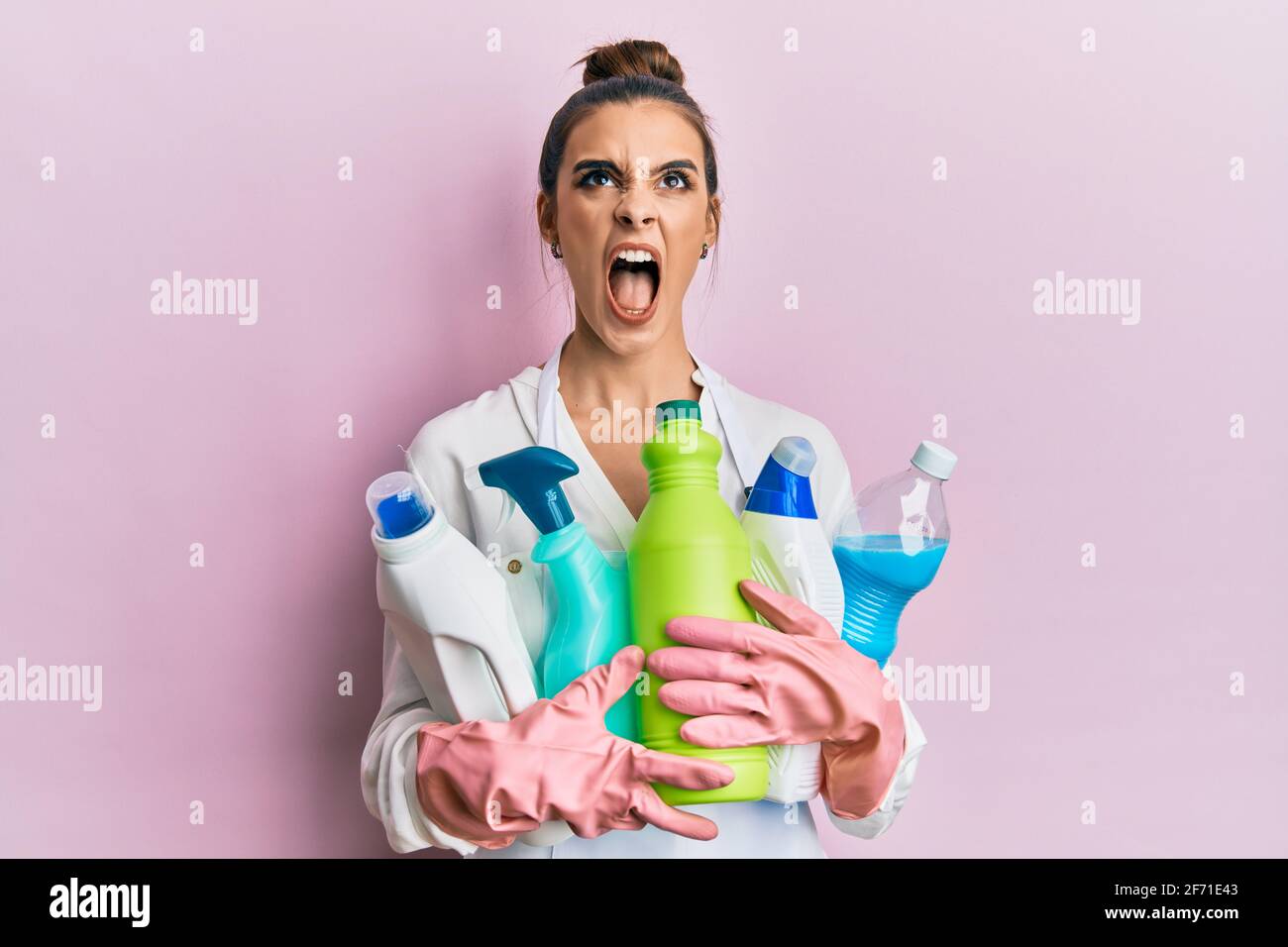 Beautiful brunette young woman wearing cleaner apron holding cleaning products angry and mad ...