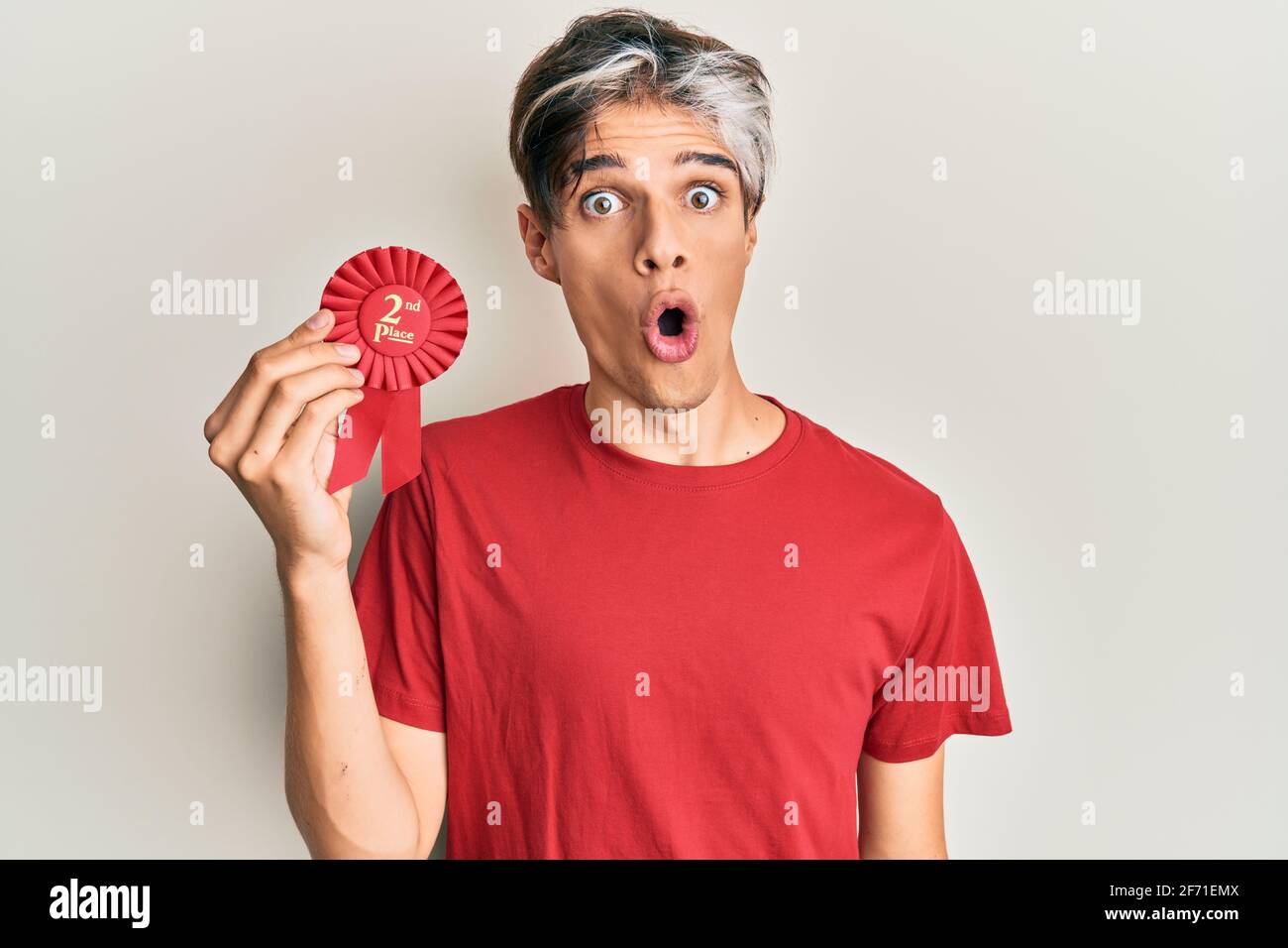 Young hispanic man holding second place badge scared and amazed with ...
