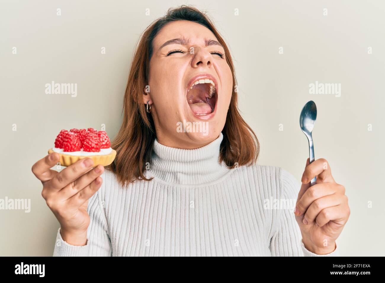 Young caucasian woman holding sweet pastry angry and mad screaming ...