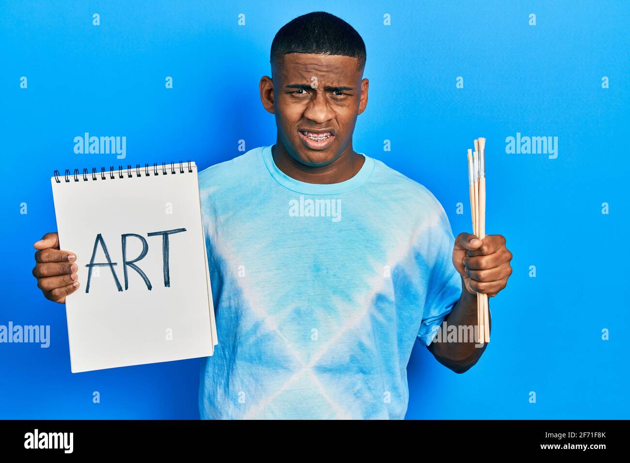 Young black man holding art notebook and painter brushes clueless and ...