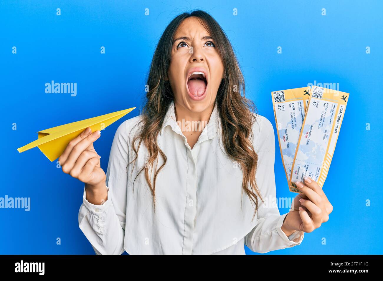Young brunette woman holding paper airplane and boarding pass angry and ...