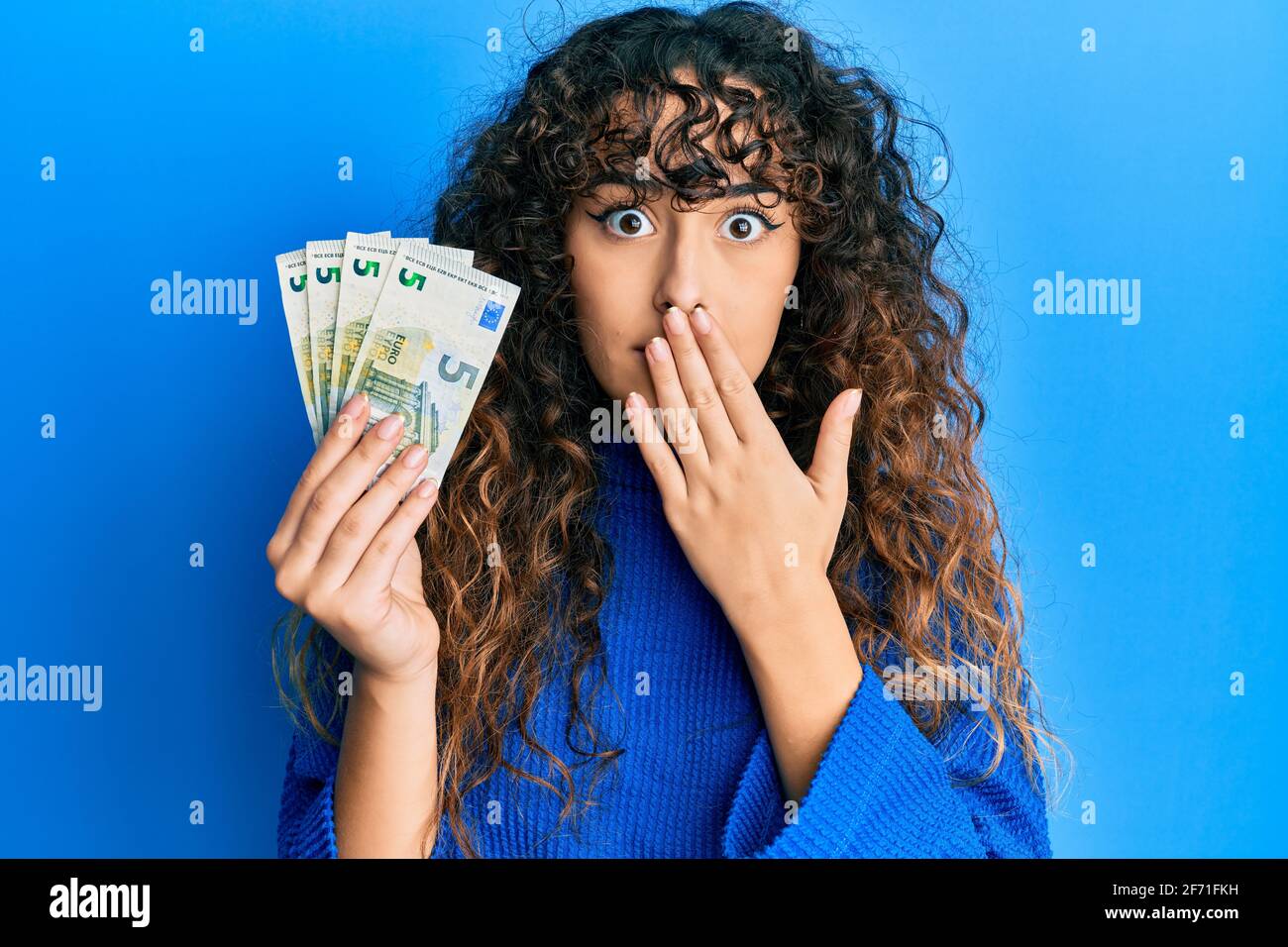 Young hispanic girl holding euro banknotes covering mouth with hand ...