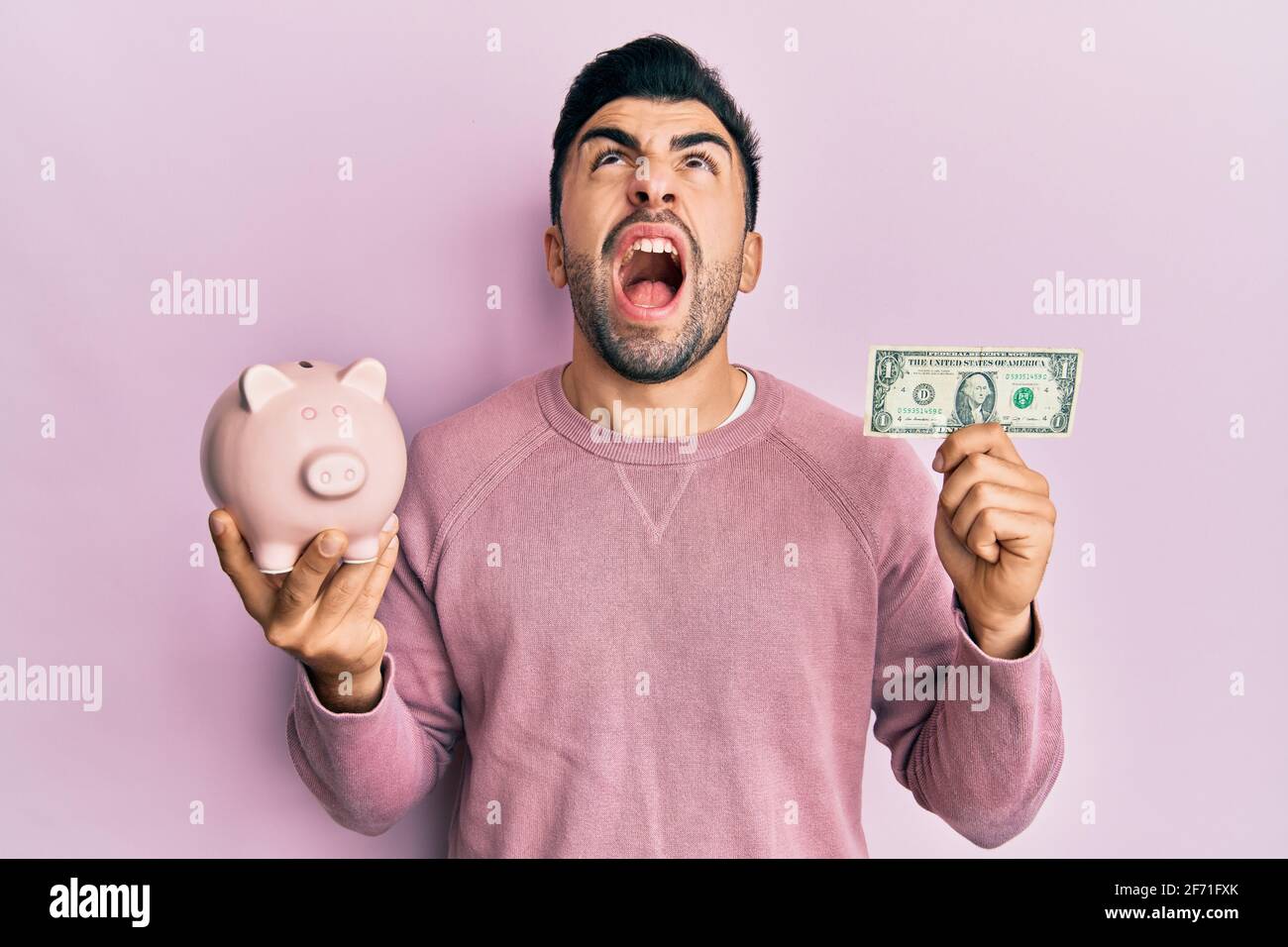 Young hispanic man holding one dollar banknote and piggy bank angry and ...