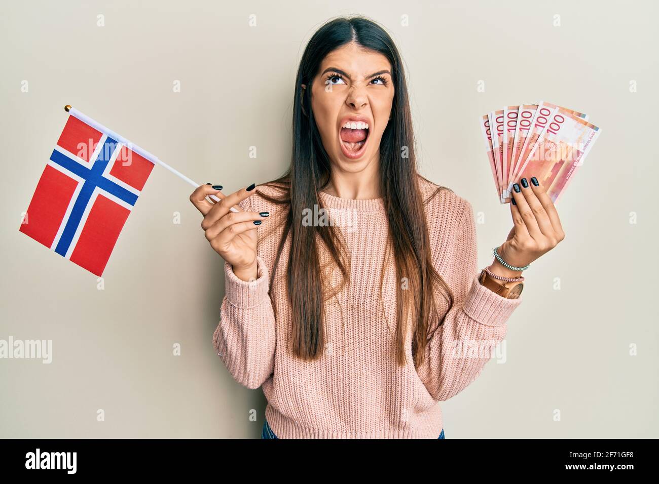 Young hispanic woman holding norway flag and krone banknotes angry and ...