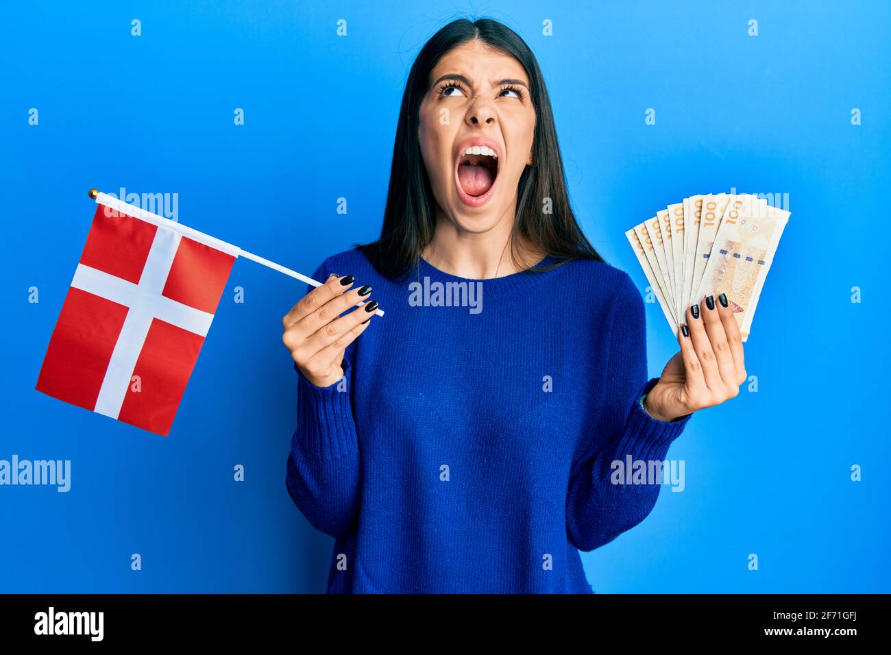 Young hispanic woman holding denmark flag and krone banknotes angry and ...