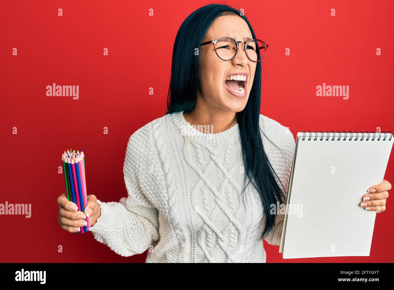 Beautiful hispanic woman holding canvas book and colored pencils angry ...