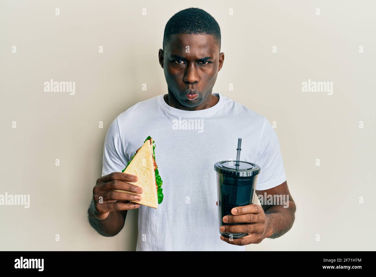 Young african american man eating sandwich and drinking soda skeptic ...