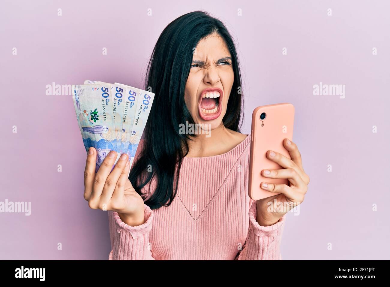 Young hispanic woman using smartphone holding colombian pesos banknotes ...