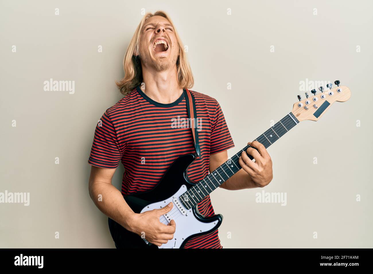Caucasian young man with long hair playing electric guitar angry and ...