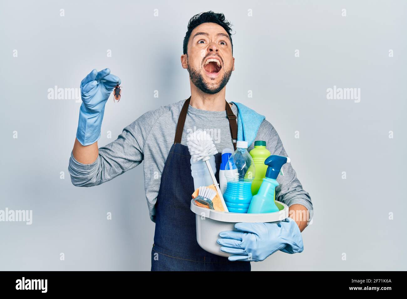 Young hispanic man holding cleaning products and cockroach angry and ...