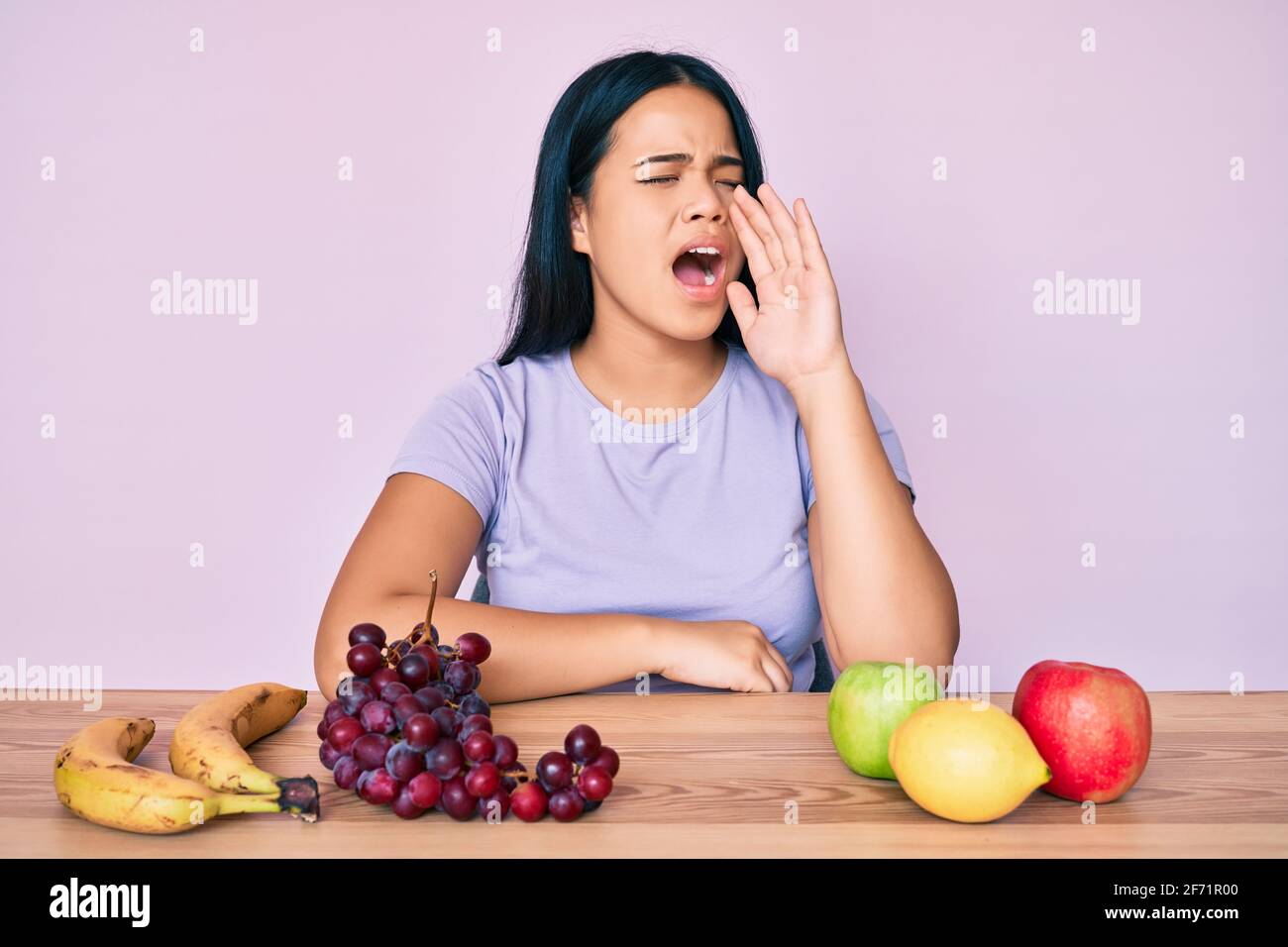 Young beautiful asian girl eating fresh and healthy fruit shouting and ...