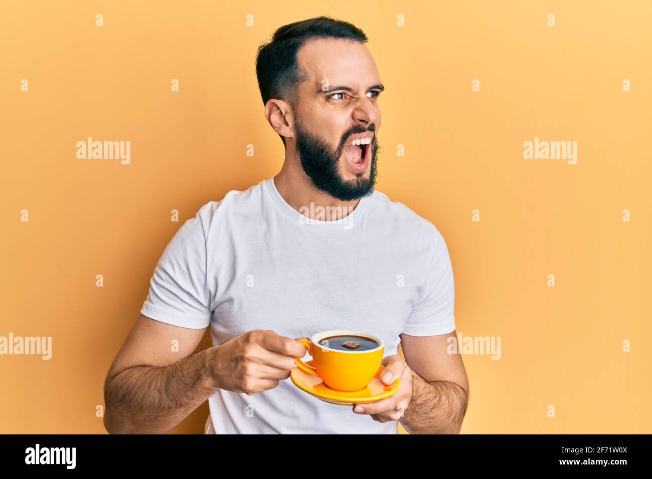 Young man with beard drinking a cup coffee angry and mad screaming ...