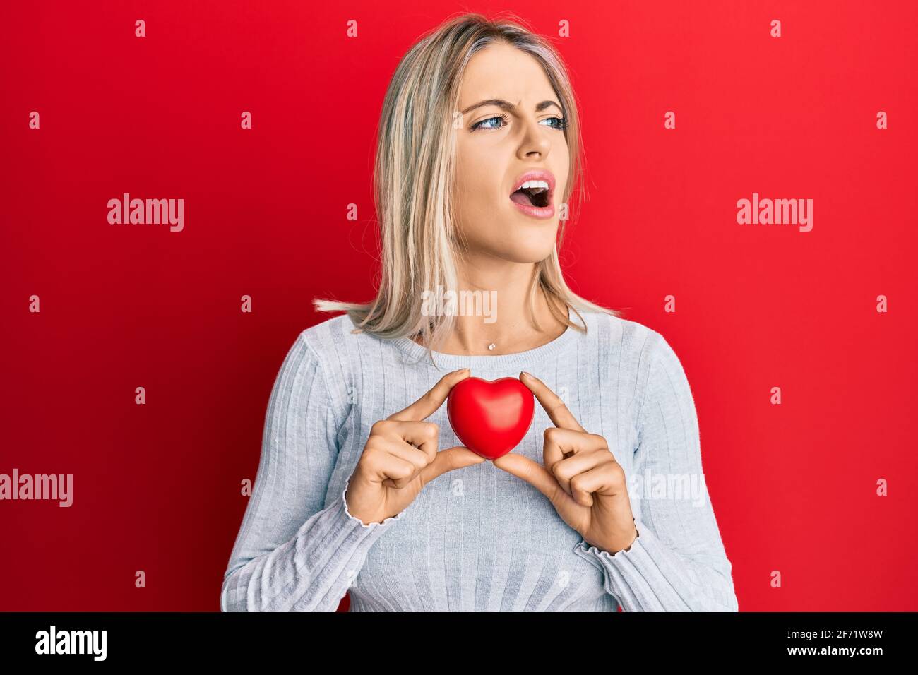 Beautiful blonde woman holding heart angry and mad screaming frustrated ...