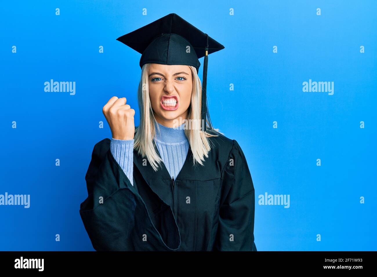 Beautiful blonde woman wearing graduation cap and ceremony robe angry ...