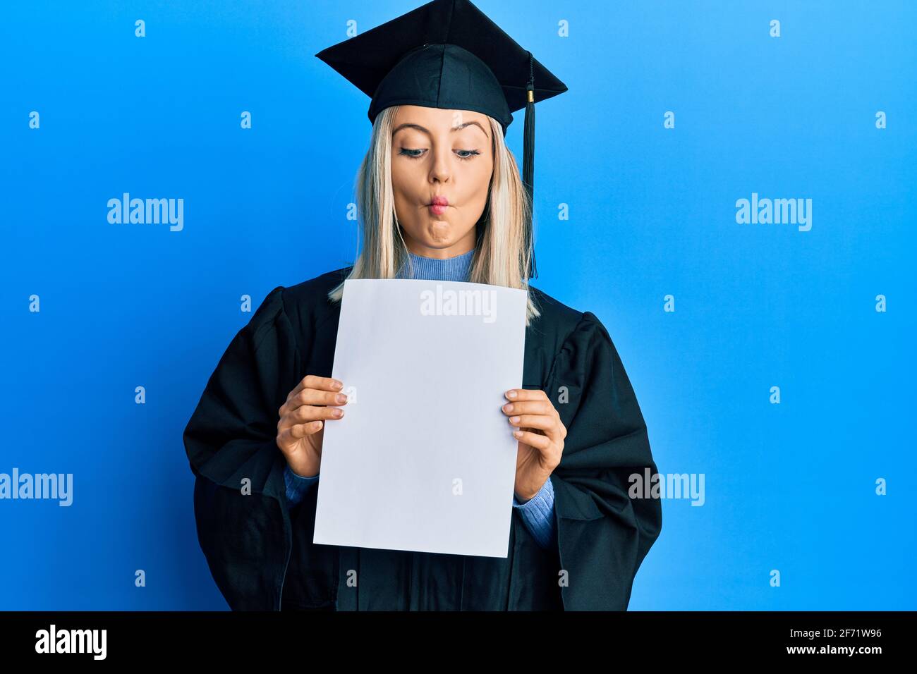 Beautiful blonde woman wearing graduation cap and ceremony robe holding ...