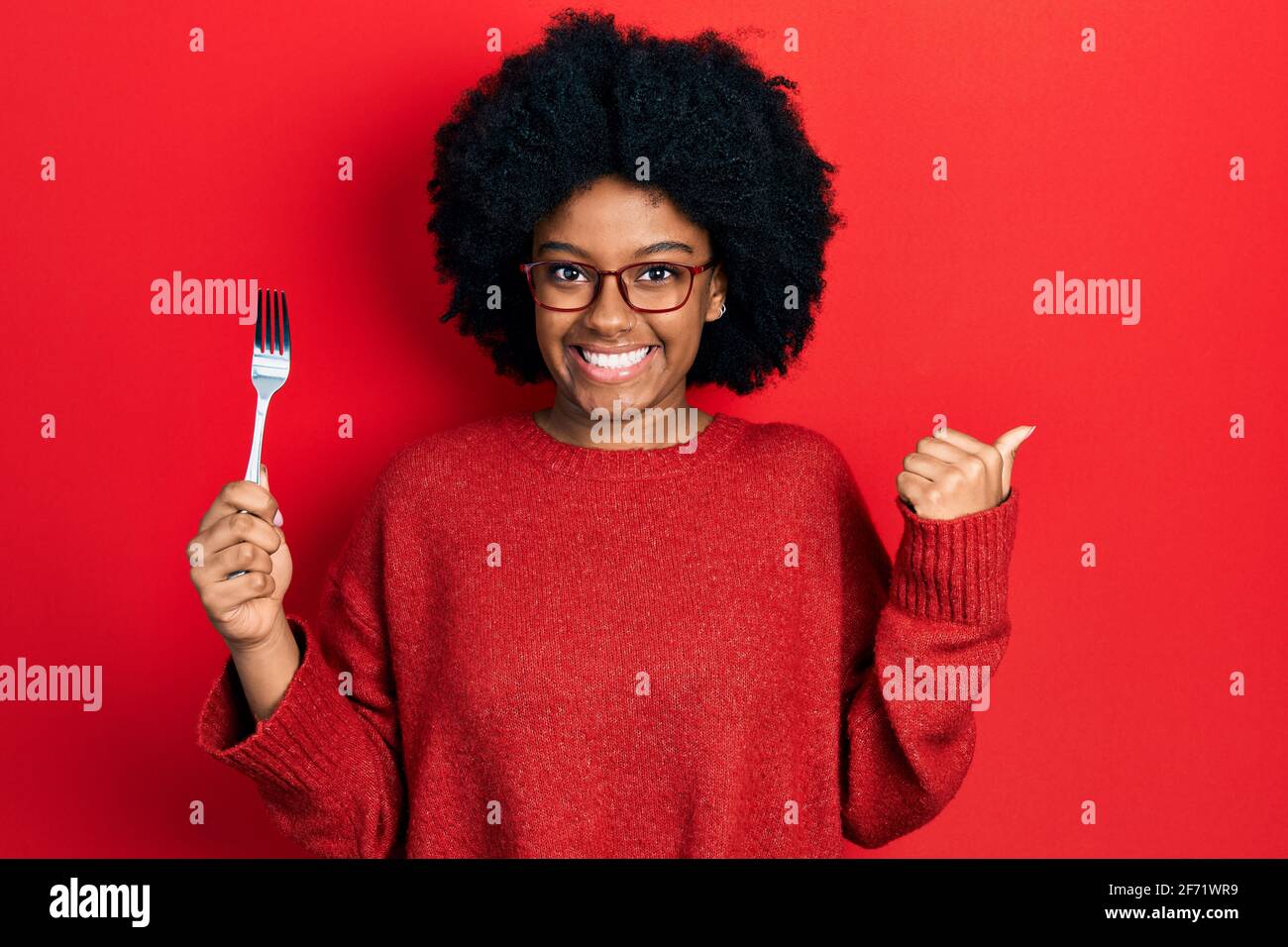 Young african american woman holding one silver fork pointing thumb up ...