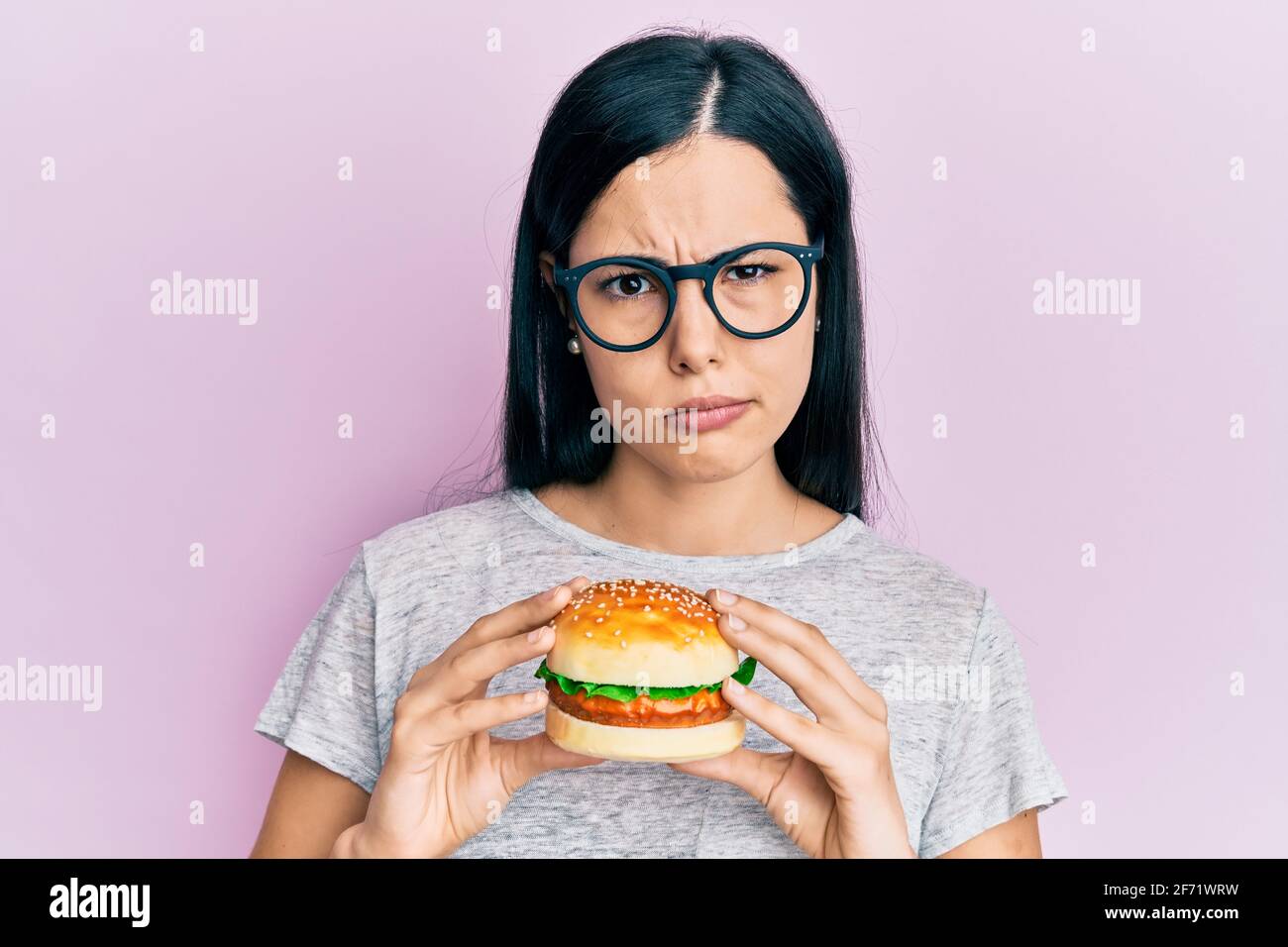 Beautiful young woman eating tasty hamburger skeptic and nervous ...