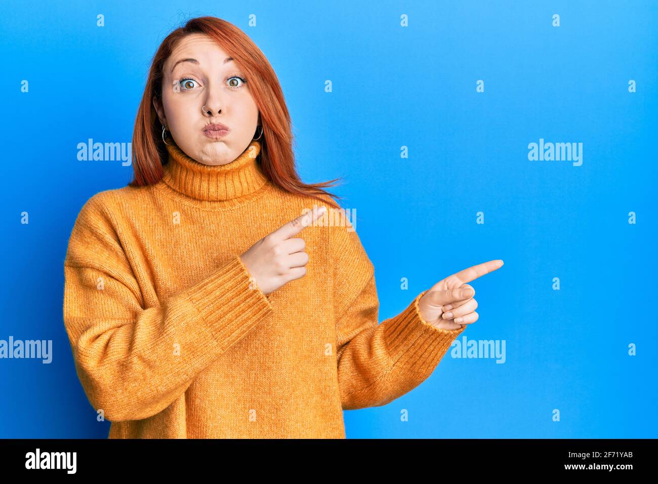 Beautiful redhead woman pointing fingers to the side puffing cheeks ...