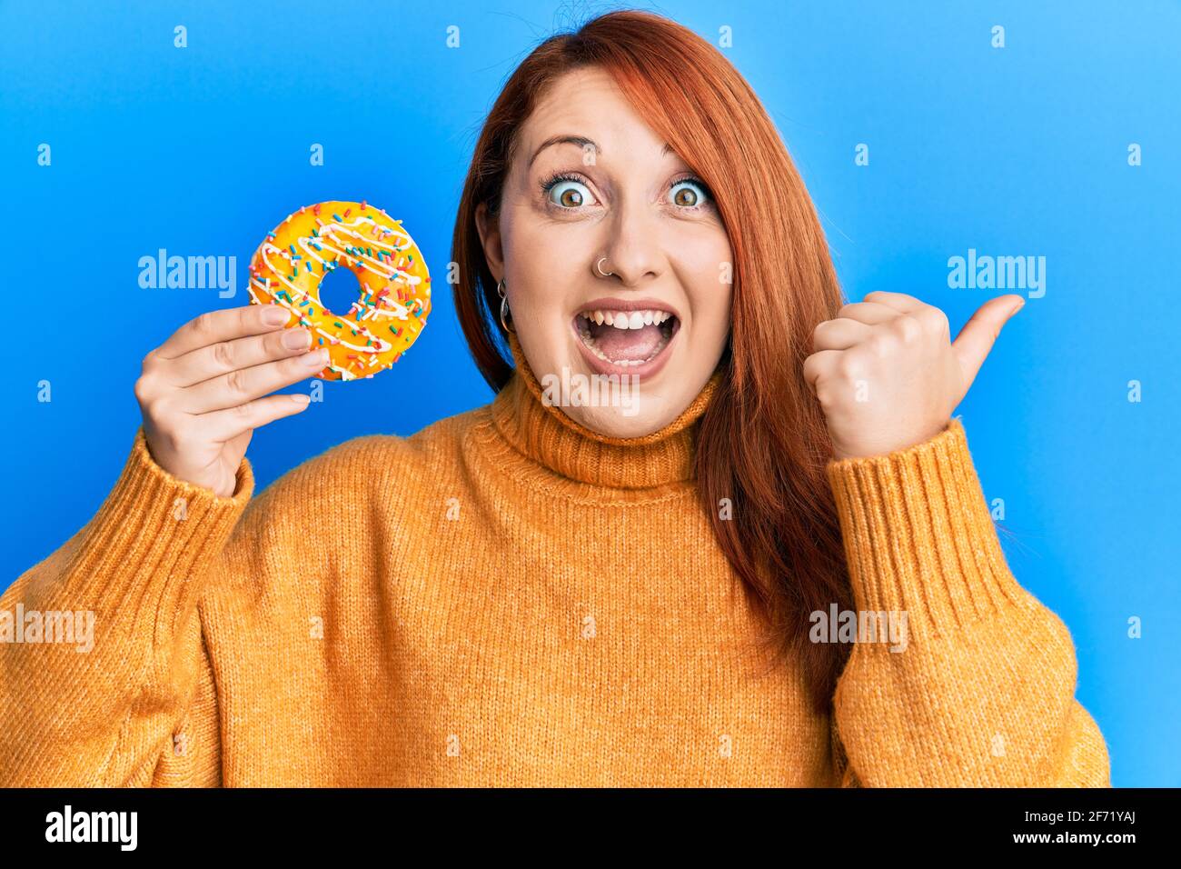 Beautiful redhead woman holding tasty colorful doughnut pointing thumb ...