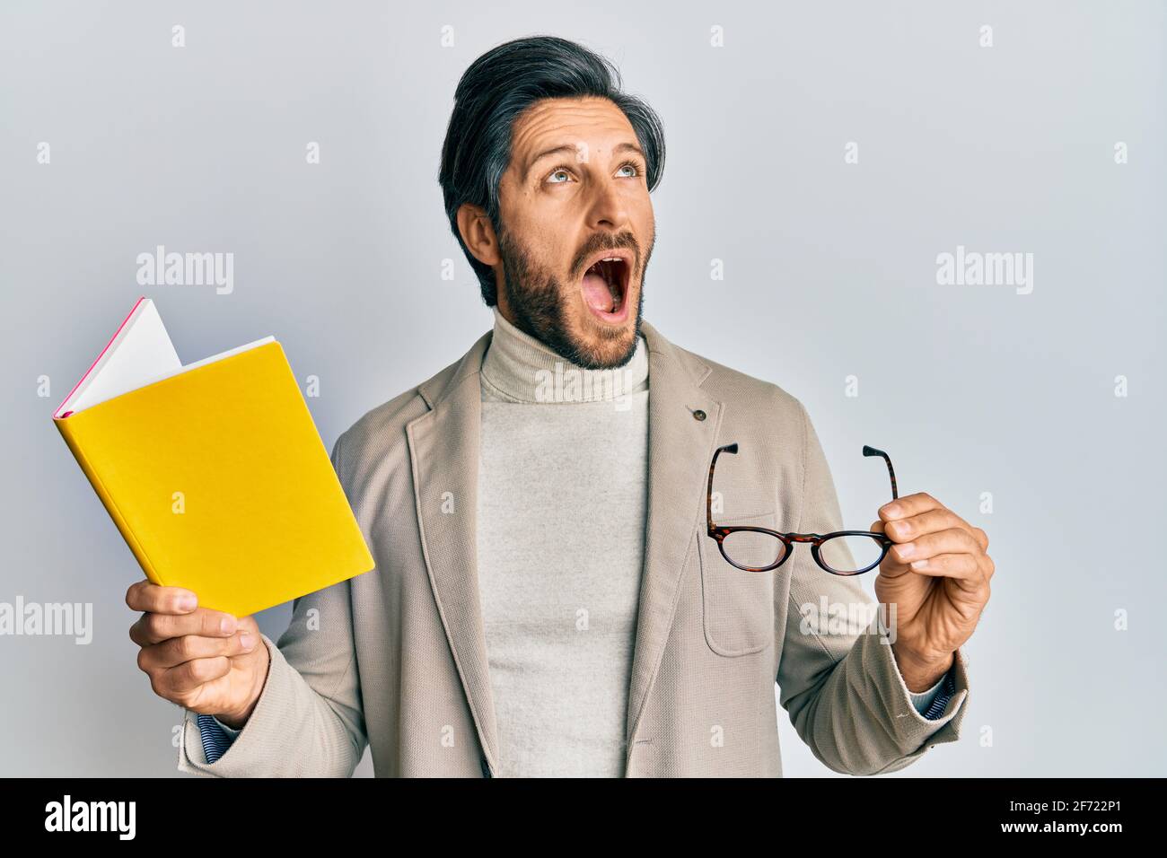 Young hispanic man reading a book and holding glasses angry and mad ...