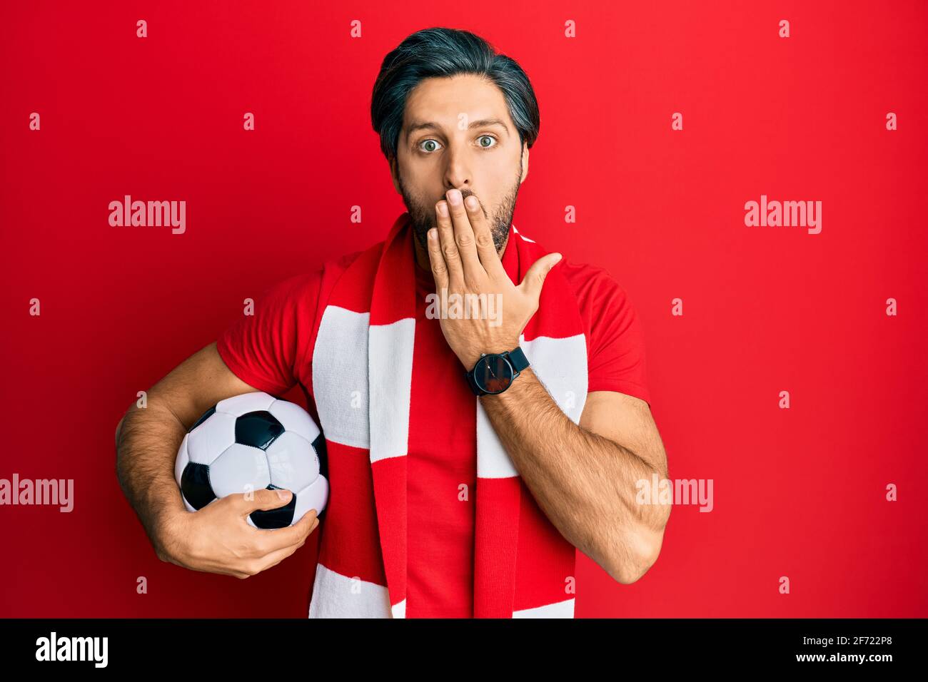 Young hispanic man football hooligan holding ball covering mouth with ...