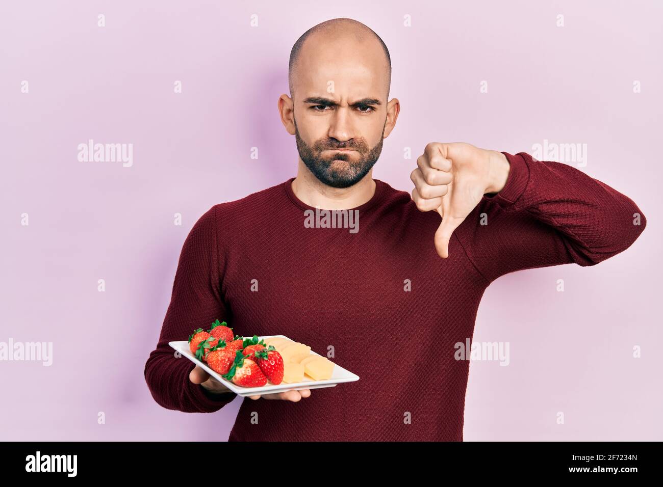 Young bald man eating fresh and healthy fruits with angry face ...