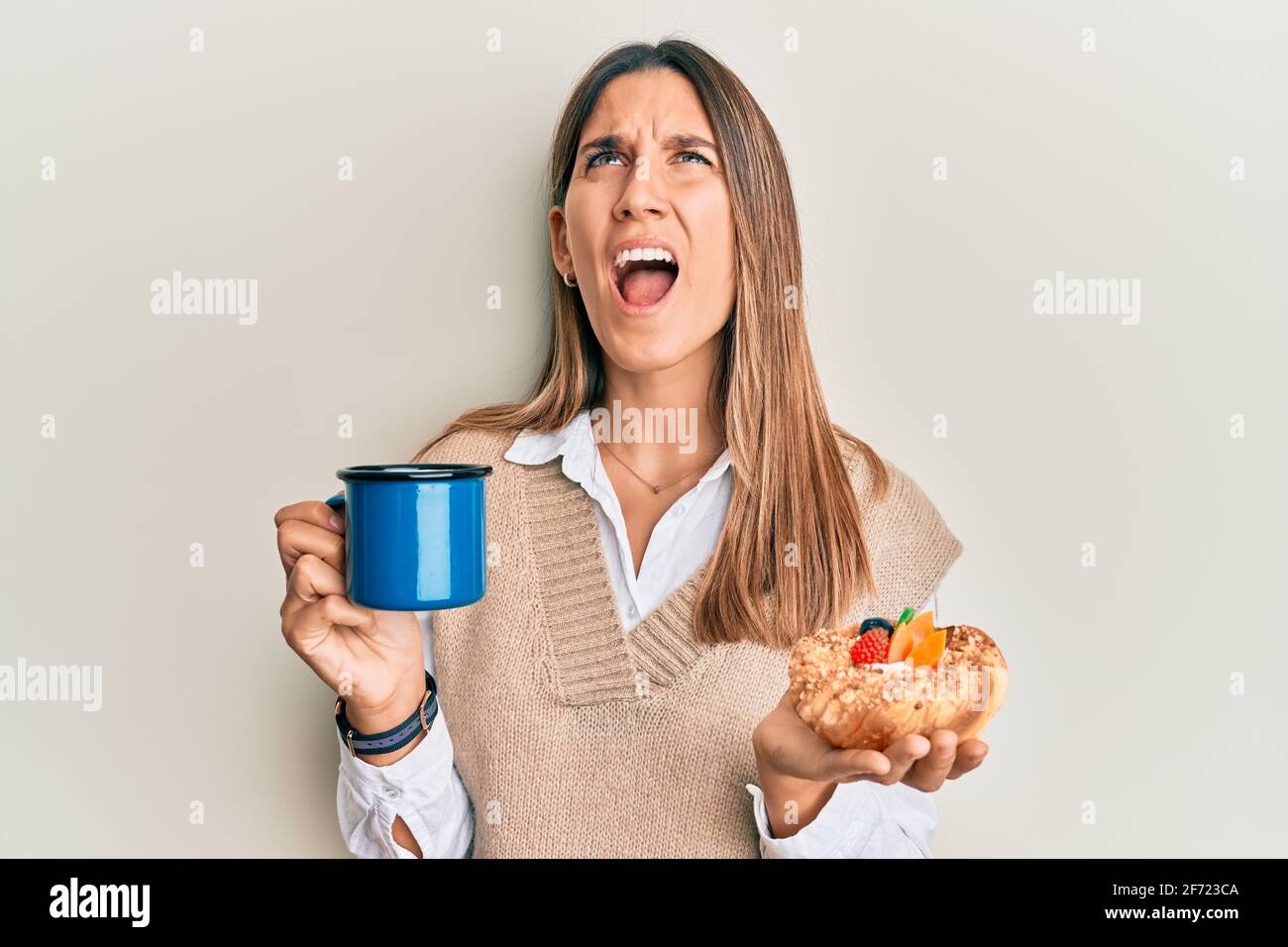 Brunette young woman drinking coffee and eating pastry angry and mad ...