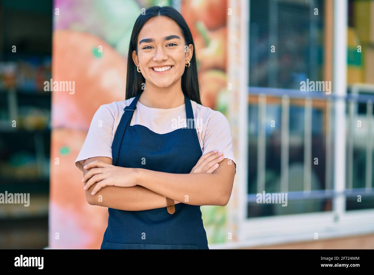 Young latin shopkeeper girl with arms crossed smiling happy at the ...
