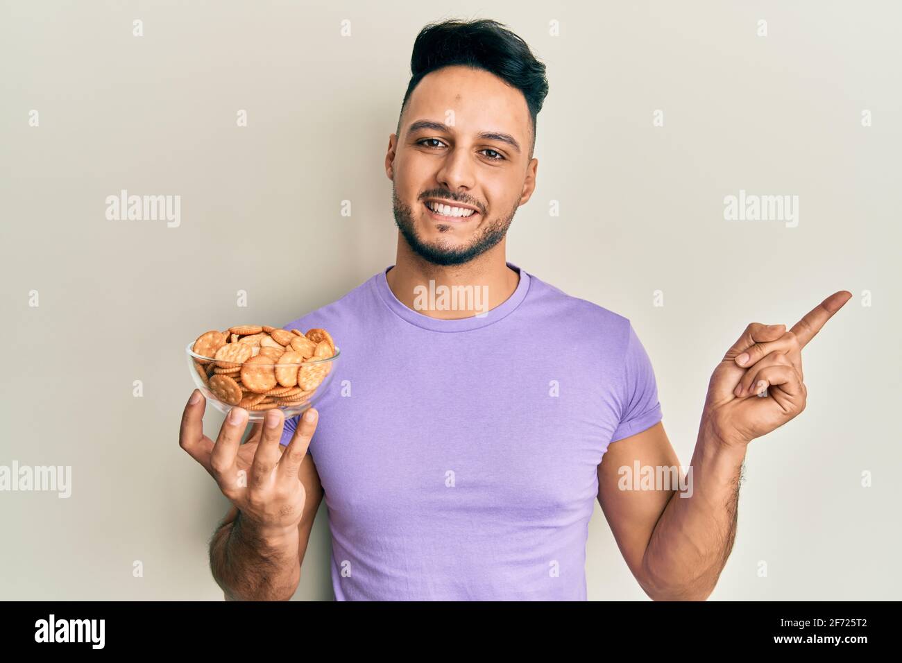 Young arab man holding bowl of salty crackers biscuits smiling happy ...