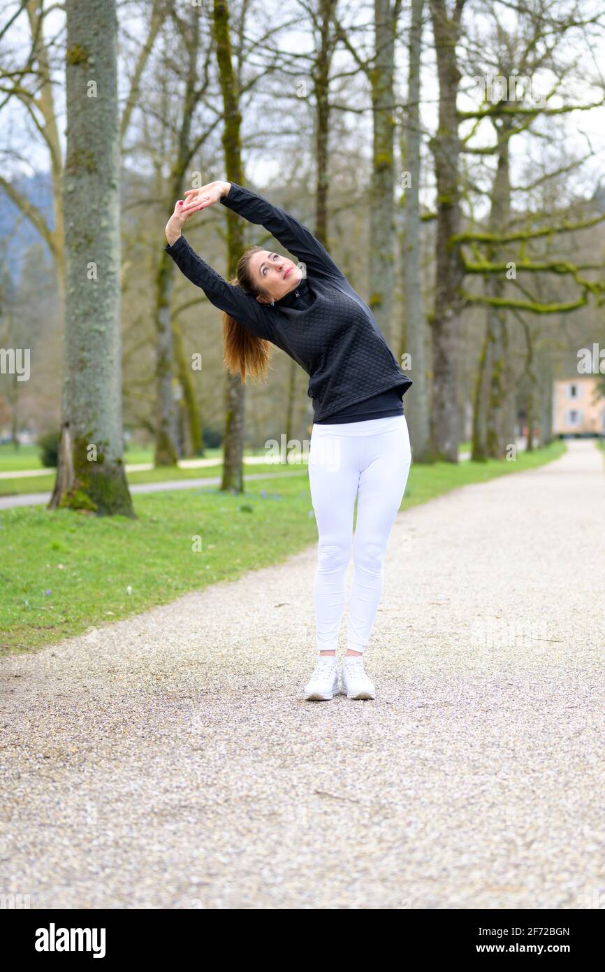 Middle-aged woman working out in a park doing stretch exercises to warm ...