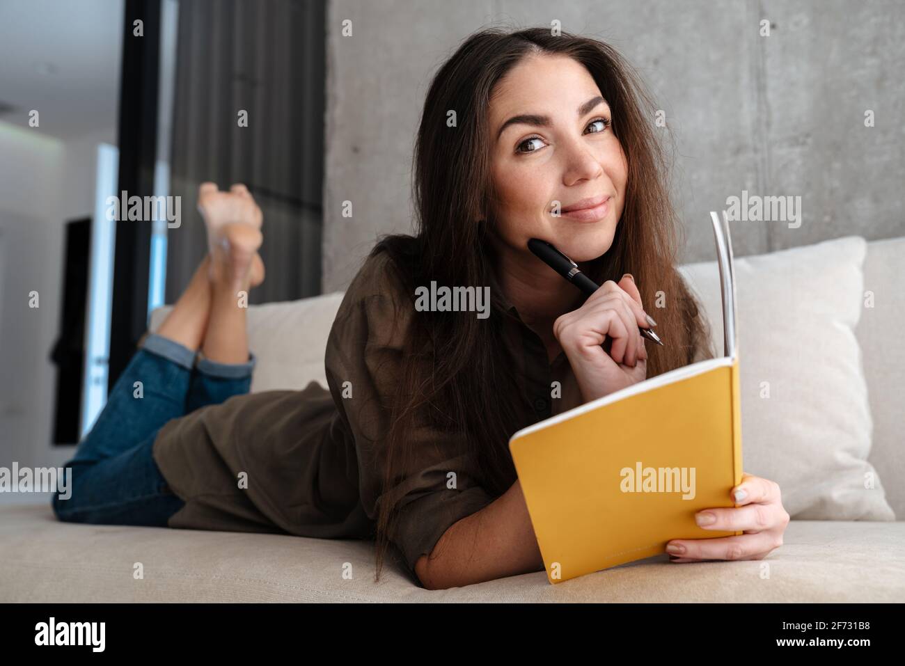 Happy brunette nice girl writing down notes while lying on sofa at home ...