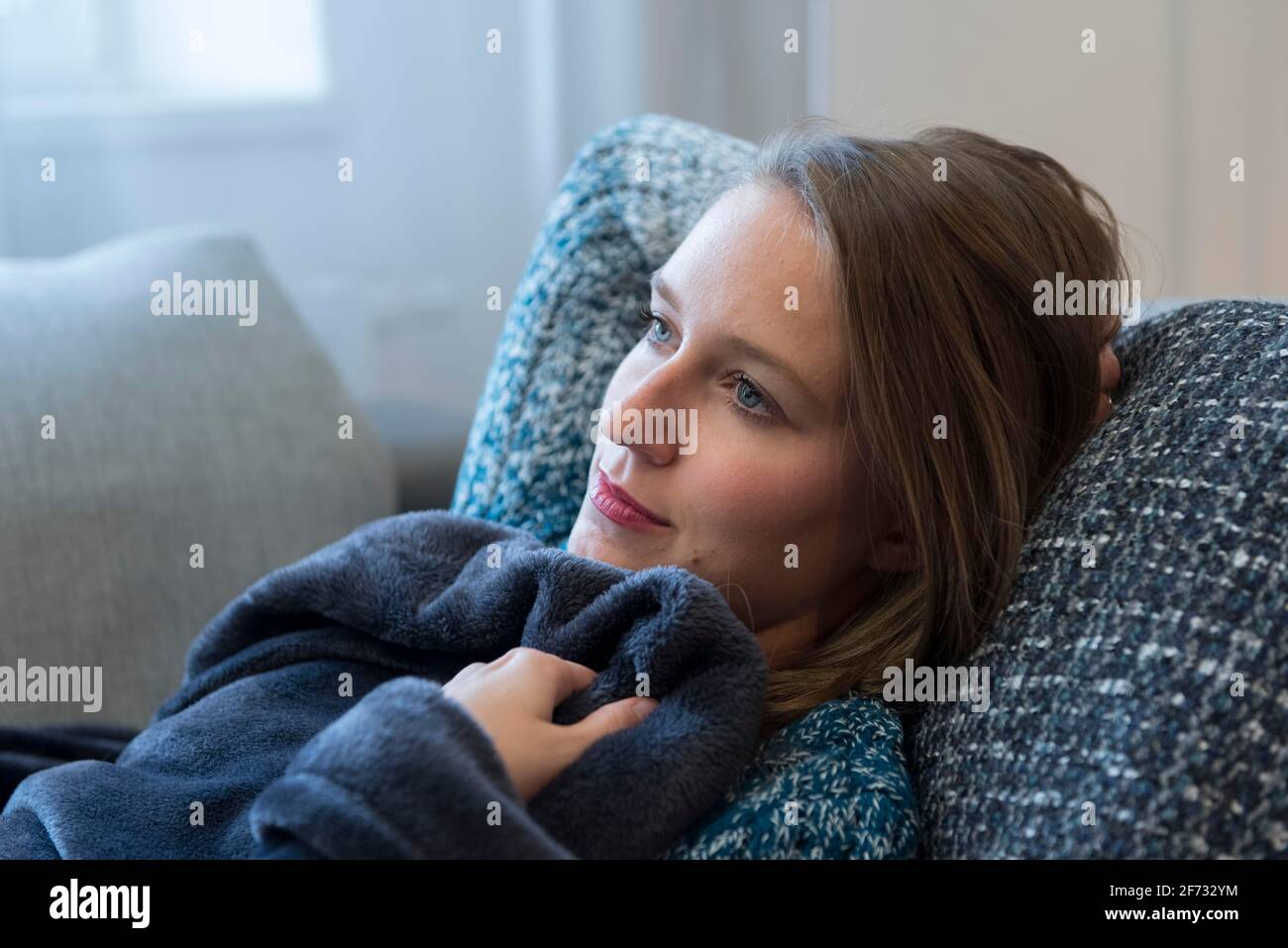 A young woman lies cuddled up in a blanket on a sofa in Dorfen, Bavaria ...
