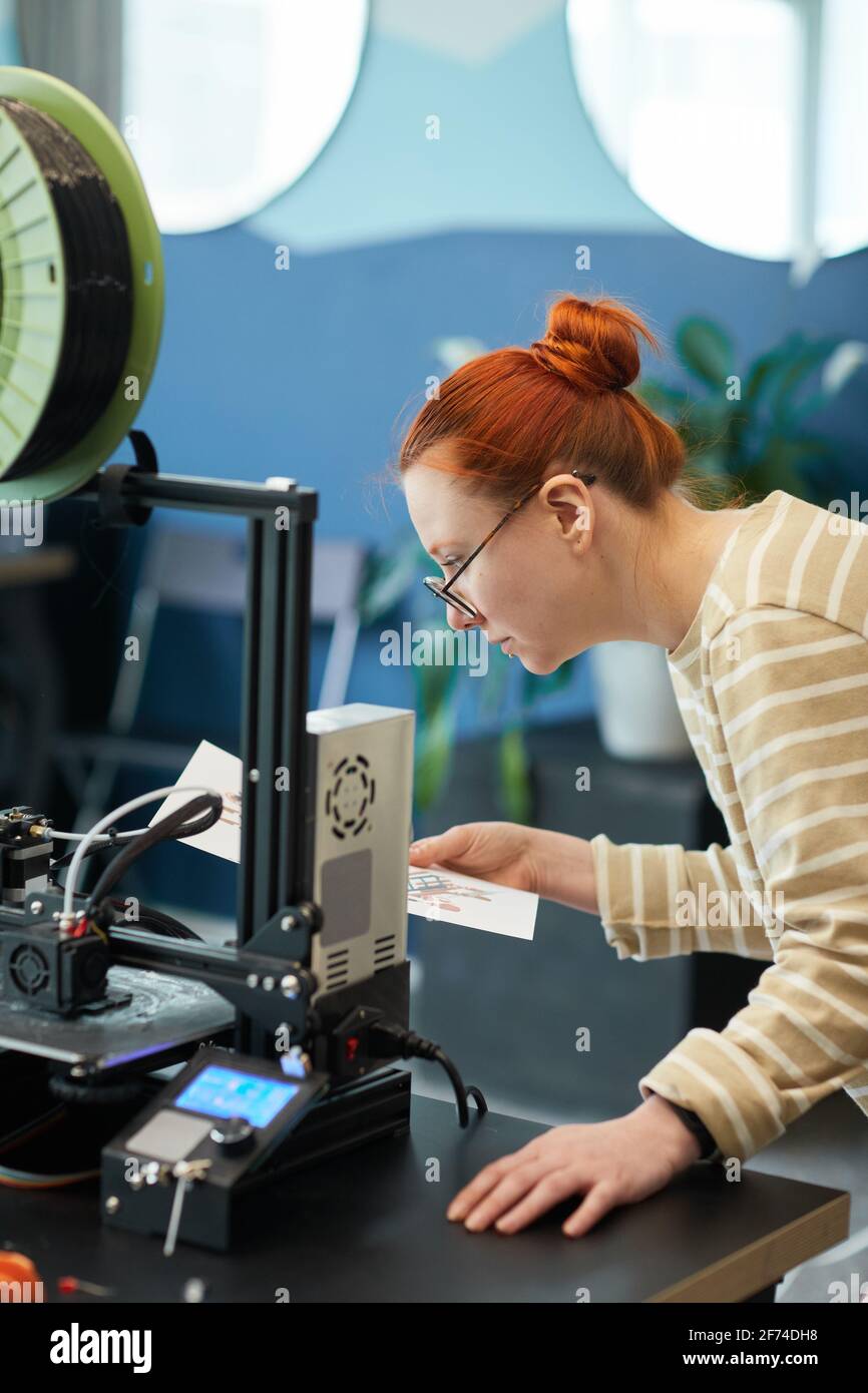 Vertical side view portrait of young red haired woman using 3D printer ...