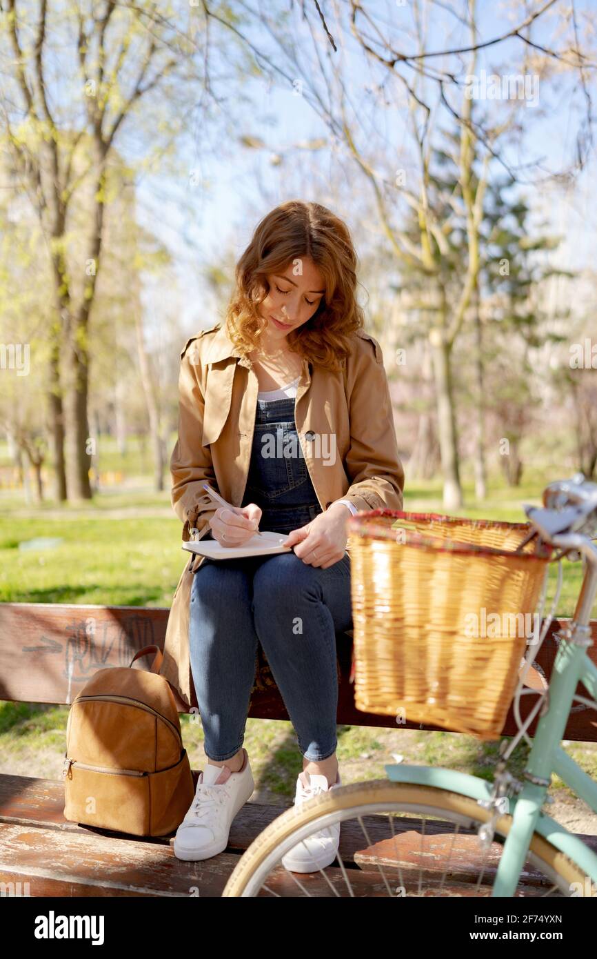 Young thoughtful female taking notes in planner on wooden bench near ...