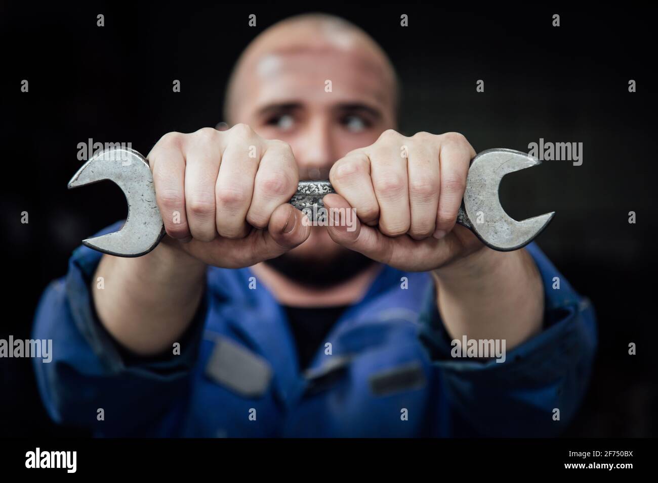 Faceless male mechanic in blue overall covering face with metal spanner ...