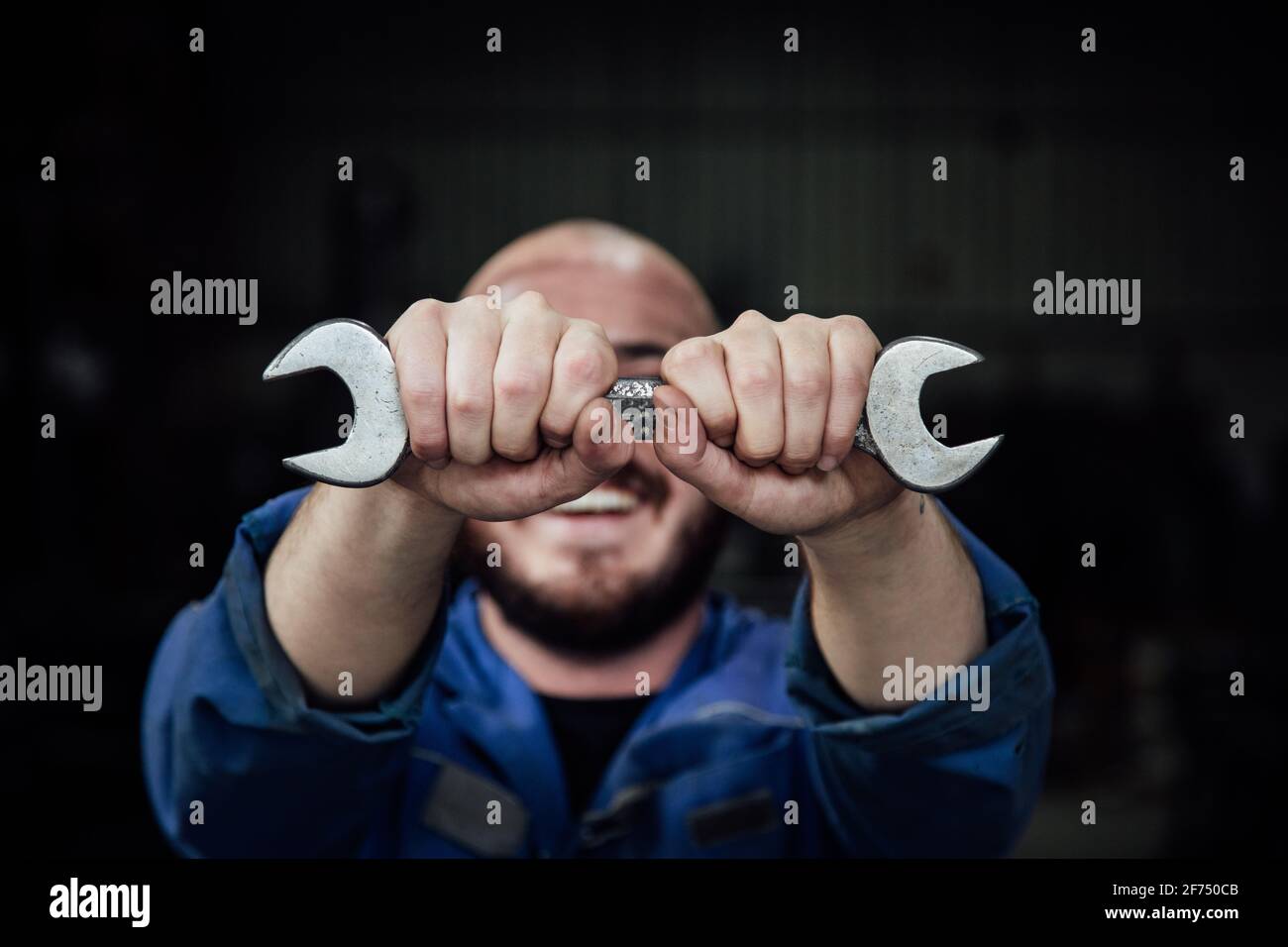 Faceless male mechanic in blue overall covering face with metal spanner ...
