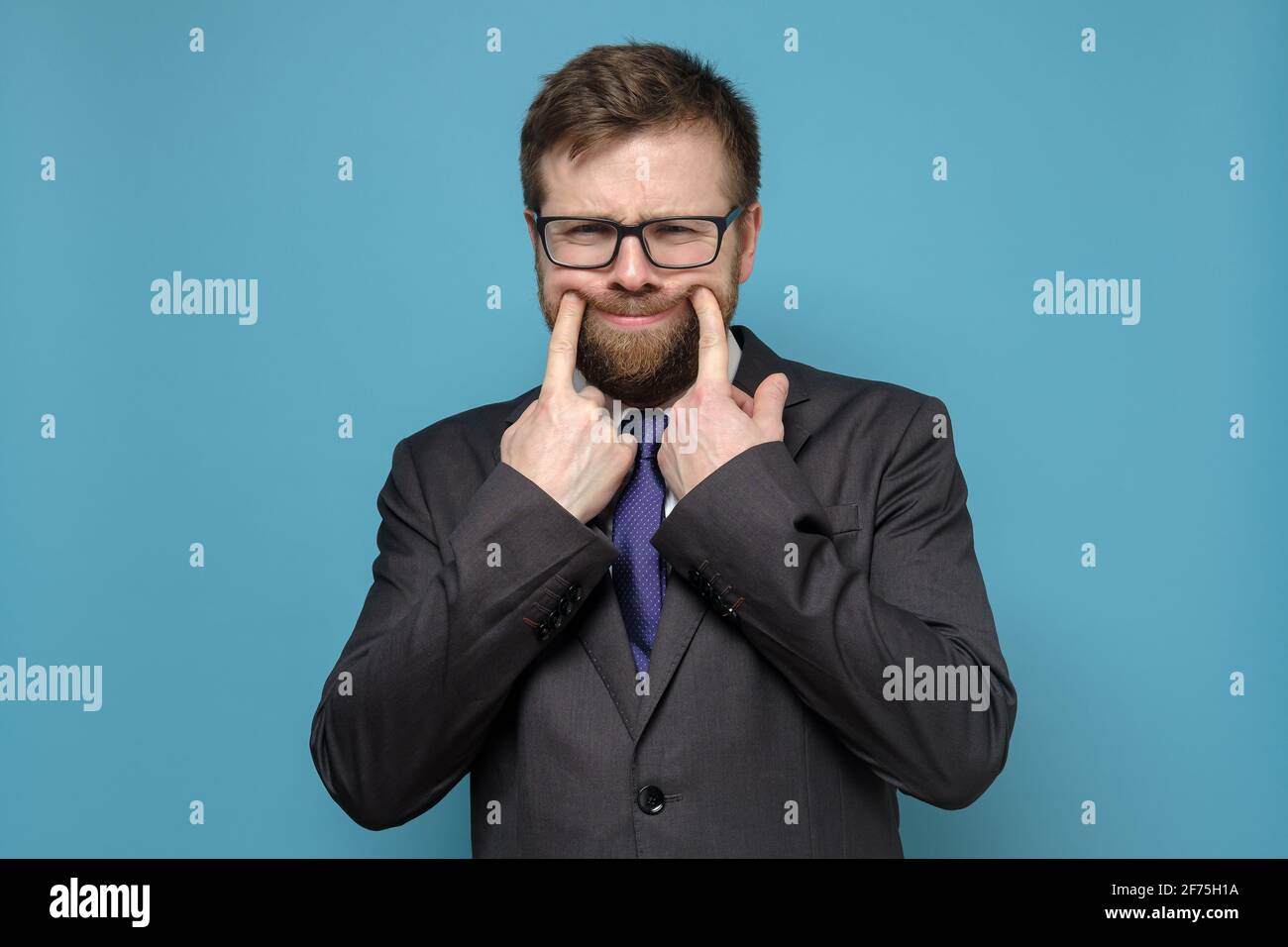 Sad man in a business suit makes a smile on his face with hands. Blue ...