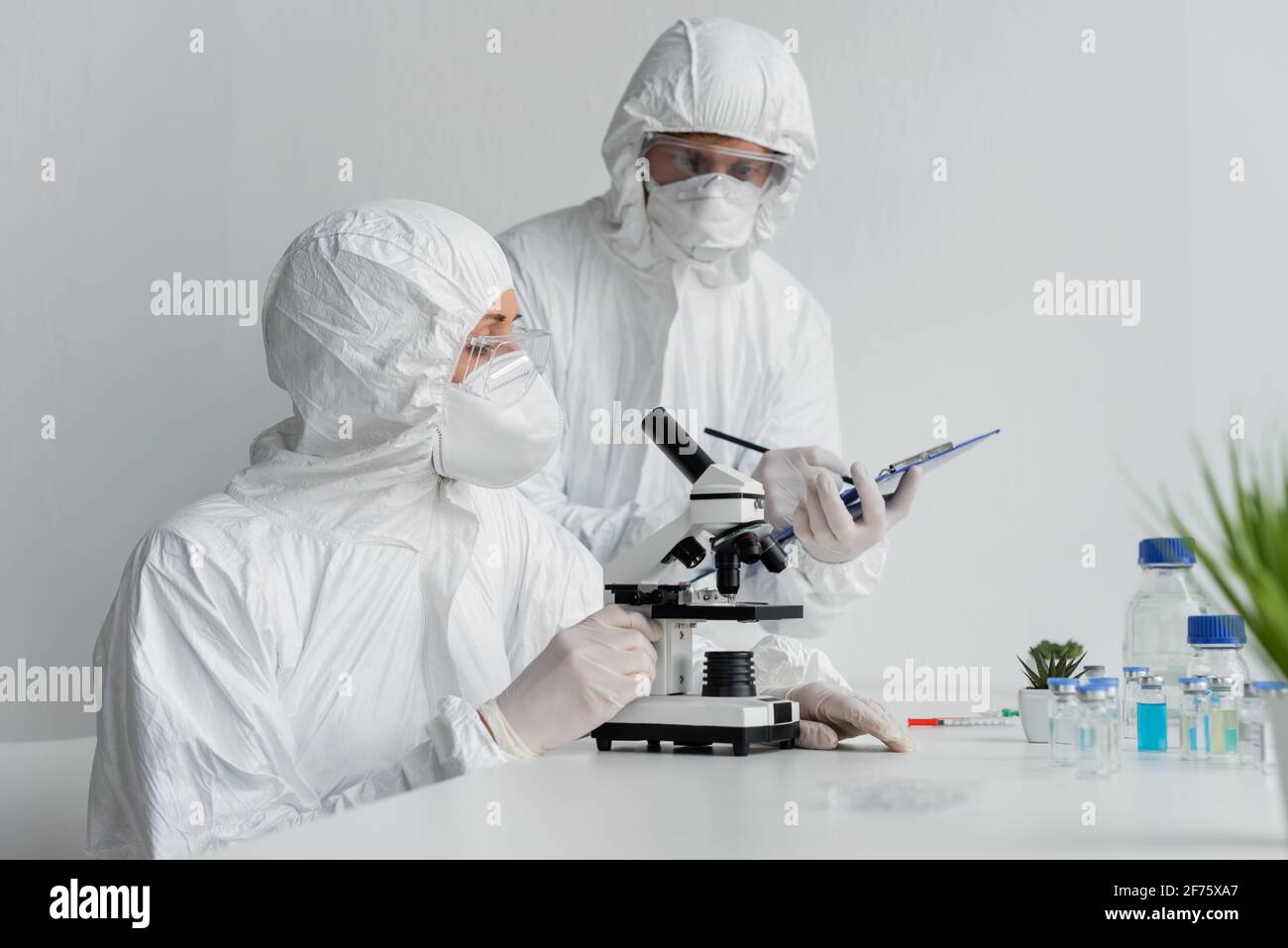 Scientist using microscope near colleague with clipboard and vaccines ...
