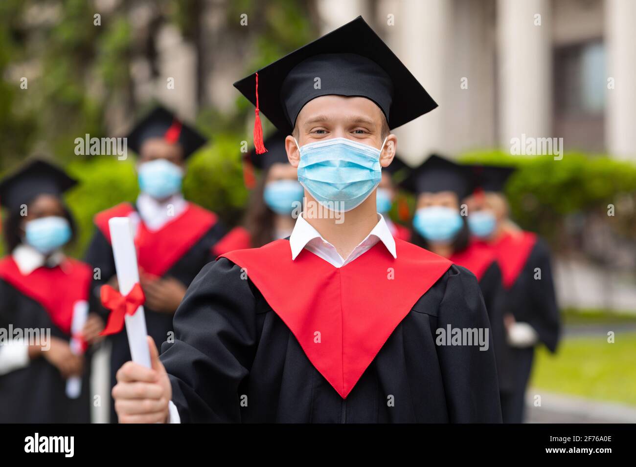 Guy student in graduation dress showing diploma, wearing face mask ...