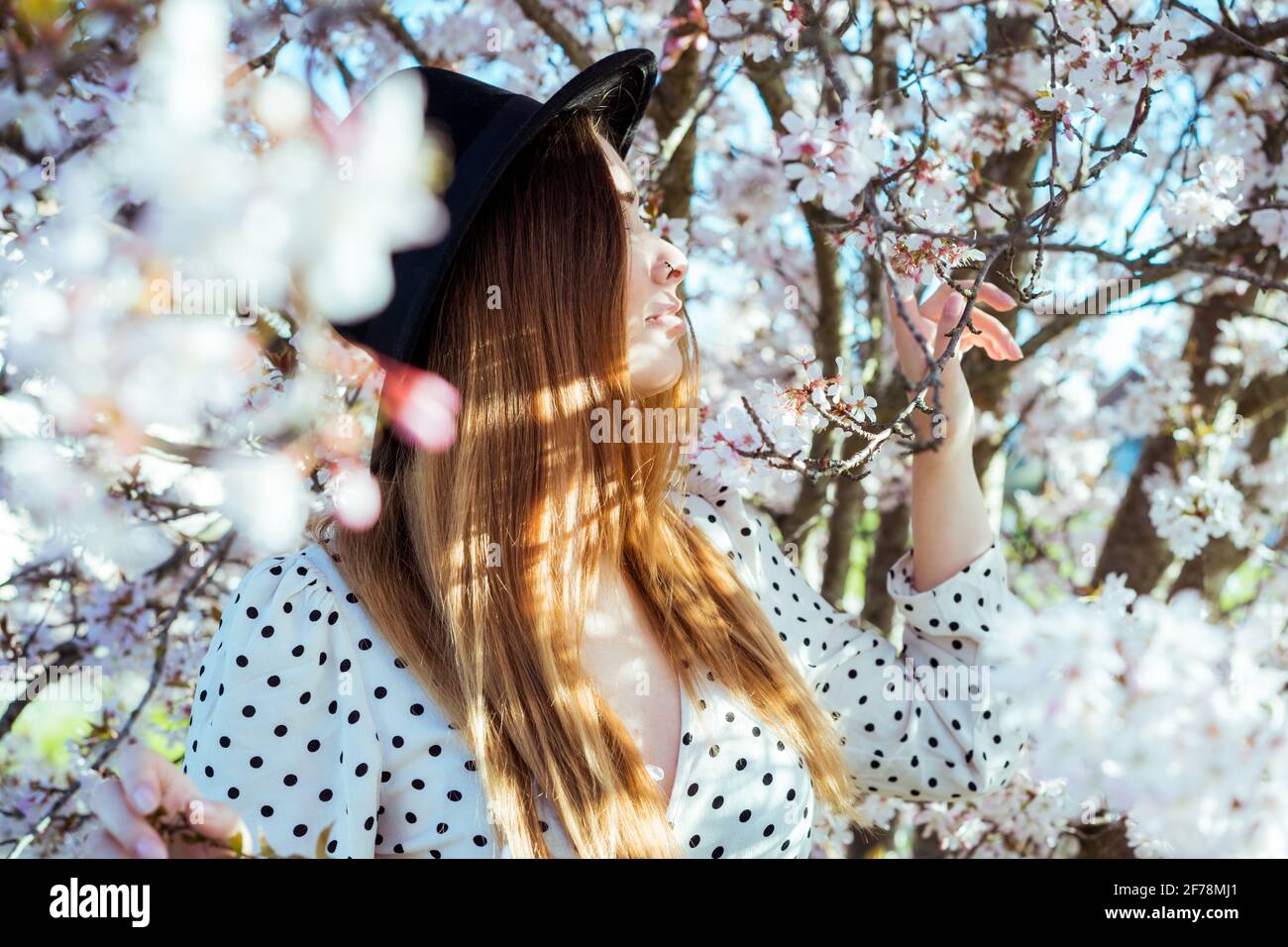 Young brunette woman in hat and dress near a white blooming tree ...