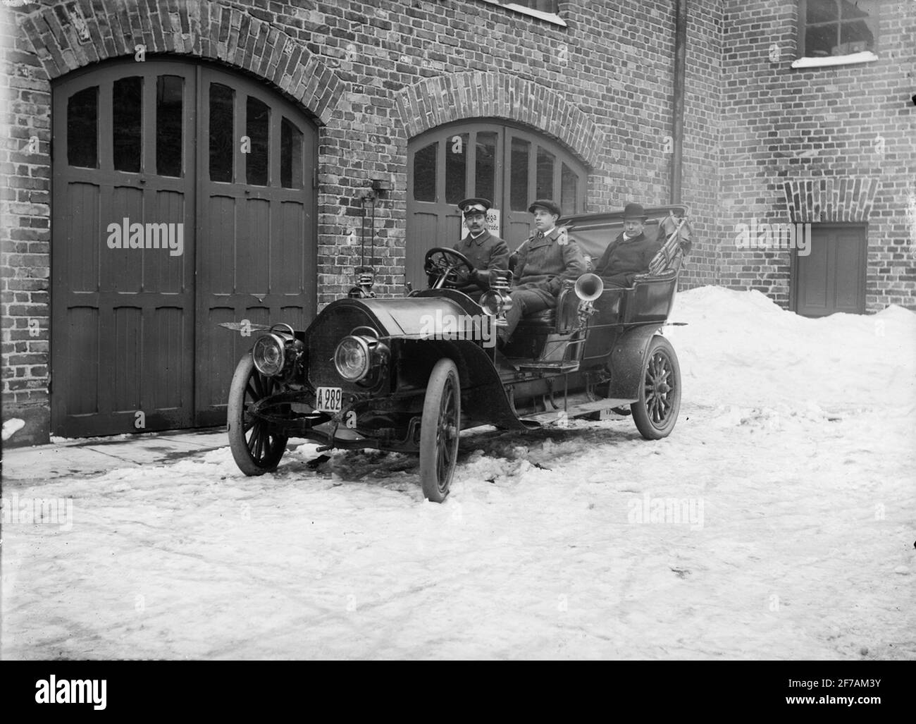 Two men with driver in car outside automobil / cart factory. Probably ...
